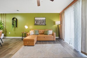 Uma sala de estar com sofá seccional, uma luminária, uma imagem emoldurada de um horizonte urbano e um vaso de planta.