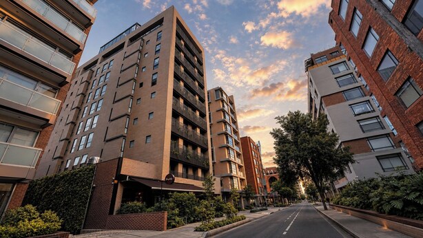 Edifício residencial de vários andares com varandas, fachada de tijolos e vista para a rua com árvores e céu claro.