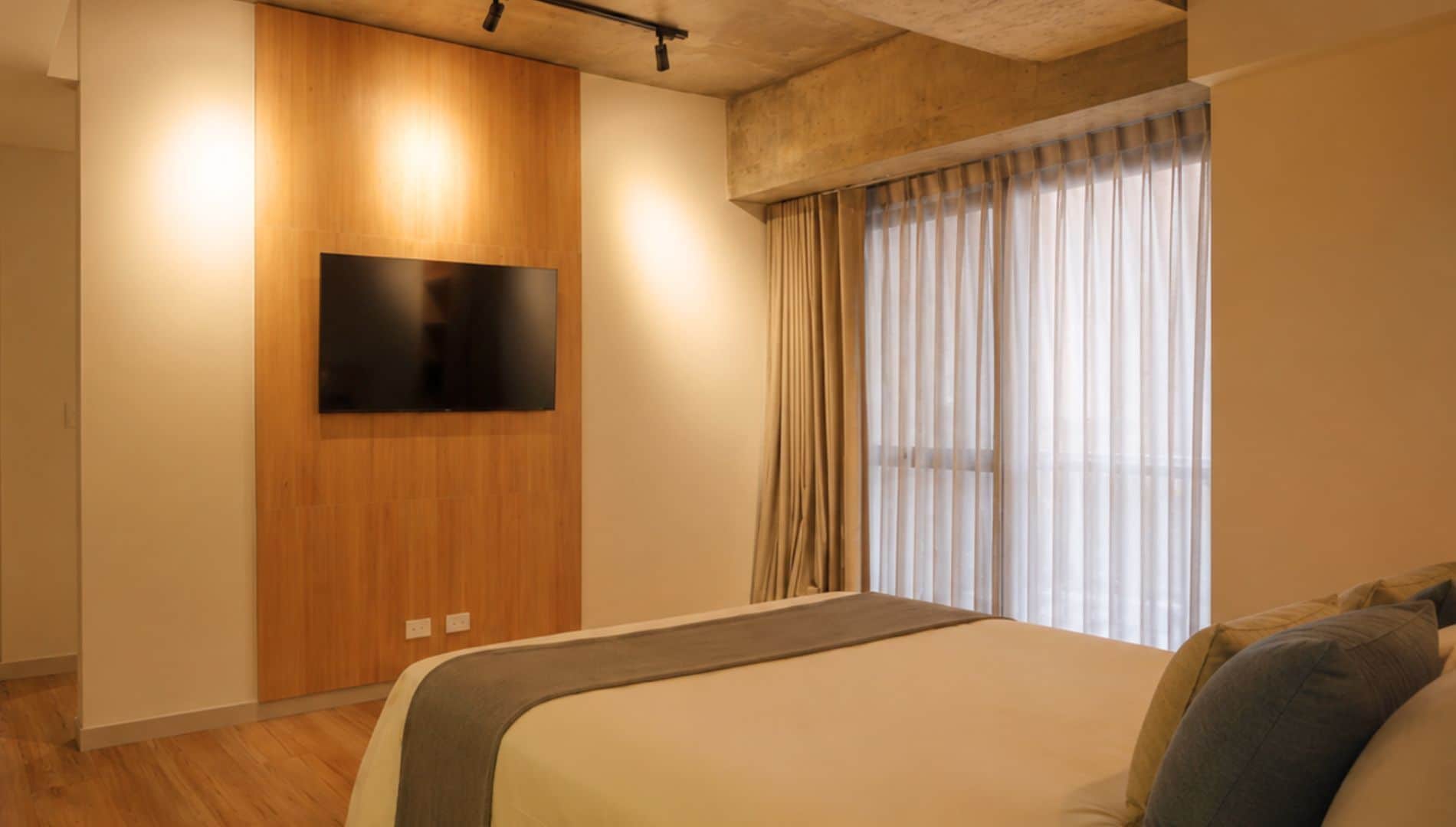 Quarto de hotel moderno com uma cama grande, televisão de tela plana na parede e janela com cortinas leves.