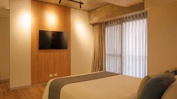 Quarto de hotel moderno com uma cama grande, televisão de tela plana na parede e janela com cortinas leves.