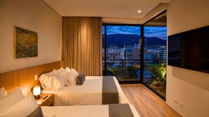 Quarto de hotel com cama, mesa de cabeceira, luminária e uma janela grande com vista para a cidade à noite.
