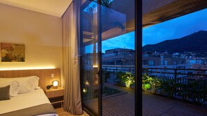 Quarto de hotel com cama, mesa de cabeceira, luminária e uma janela grande com vista para a paisagem urbana e montanhas.