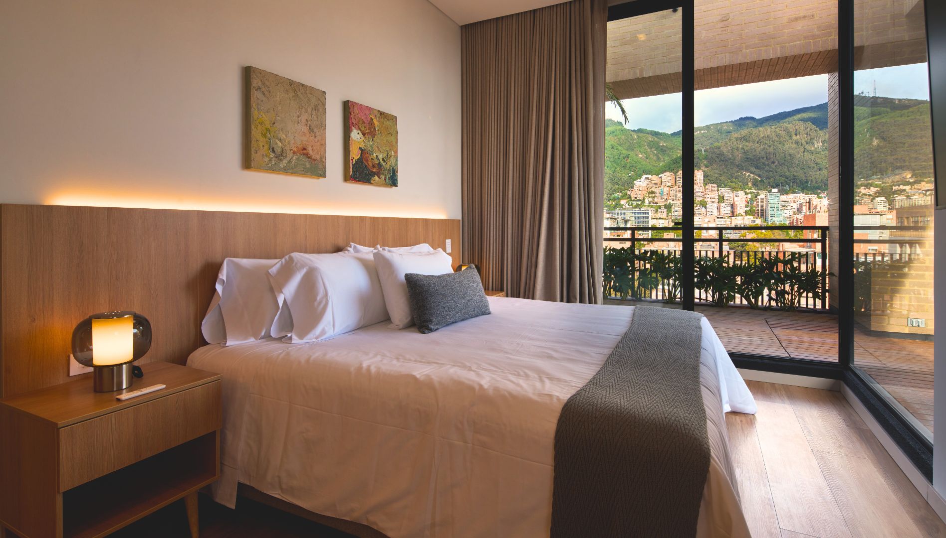 Quarto de hotel com uma cama grande, mesa de cabeceira, luminária e vista para a cidade através de uma varanda.