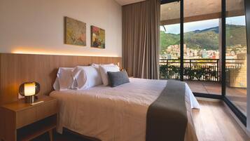 Quarto de hotel com uma cama grande, mesa de cabeceira, luminária e vista para a cidade através de uma varanda.