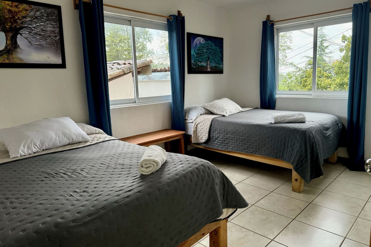 Quarto com duas camas, um banco e janelas amplas com cortinas.