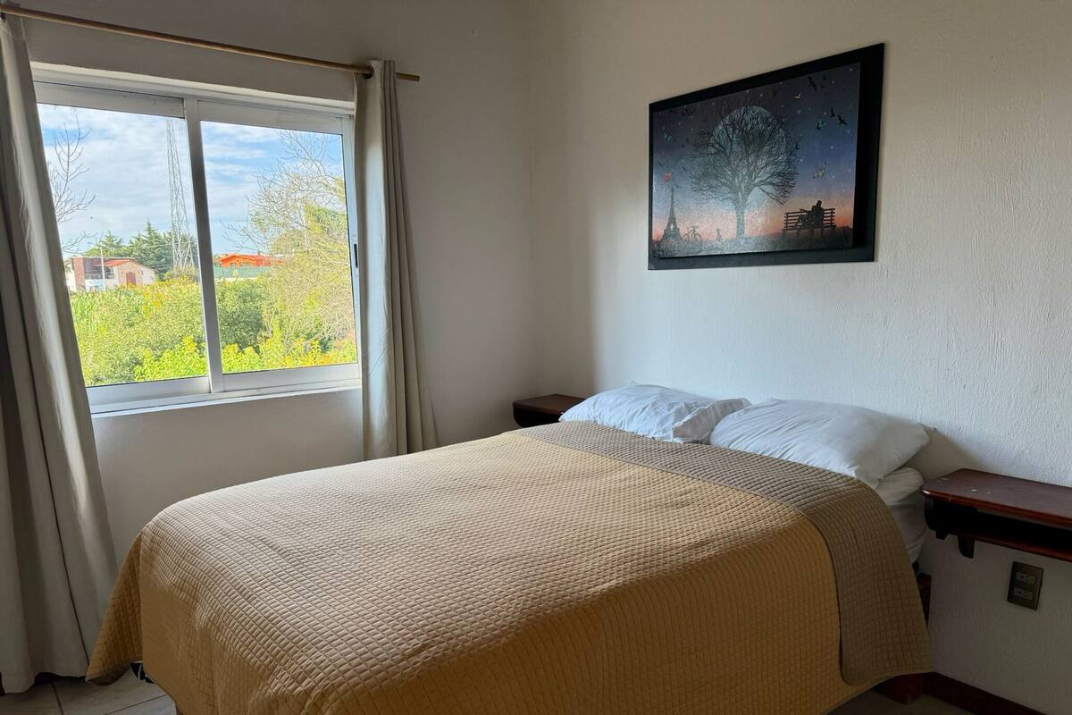 Um quarto com cama, mesa de cabeceira, janela com cortinas e um quadro na parede.