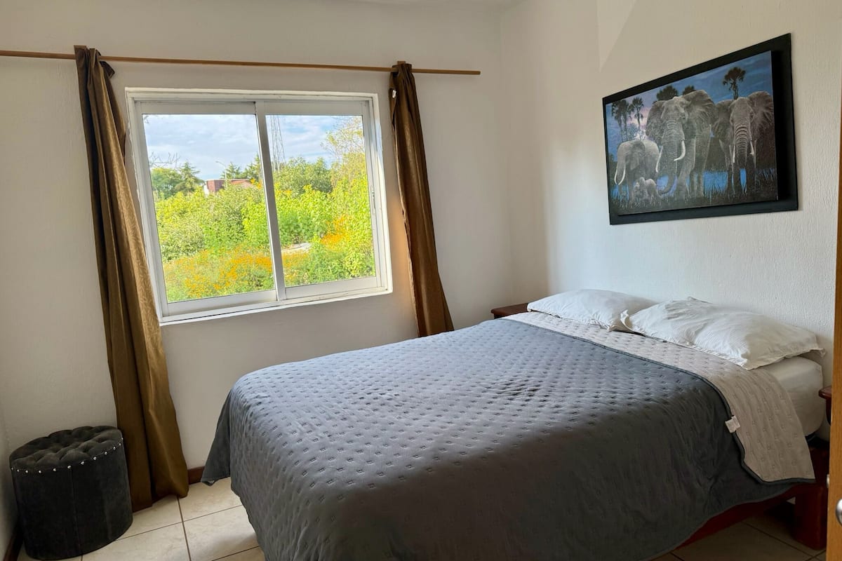 Quarto com cama, janela com cortinas, quadro na parede e uma pequena lixeira.