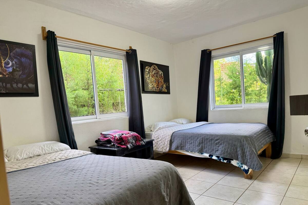 Quarto com duas camas, uma mesa pequena e uma janela com cortinas.