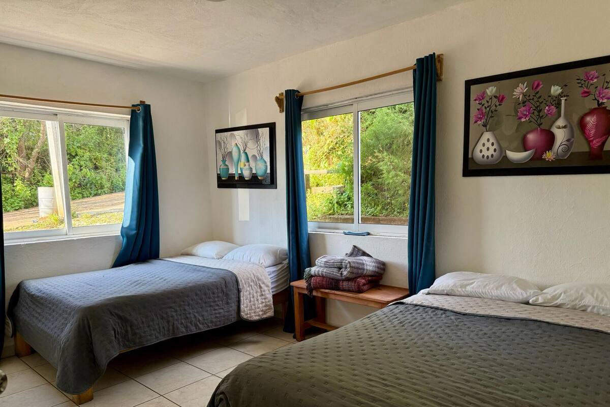 Quarto com duas camas, uma janela com cortinas, um quadro e um banco com cobertores.