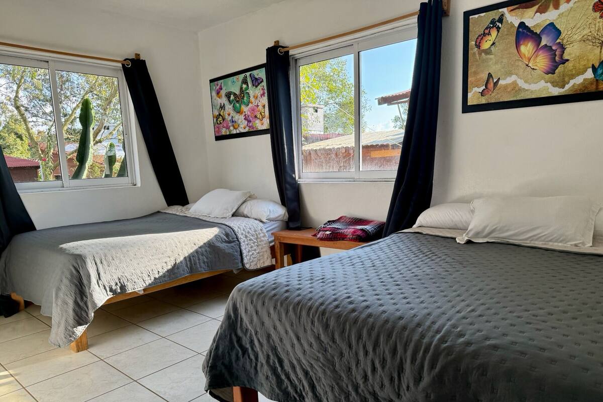 Quarto com duas camas, uma mesinha e janela com cortinas.