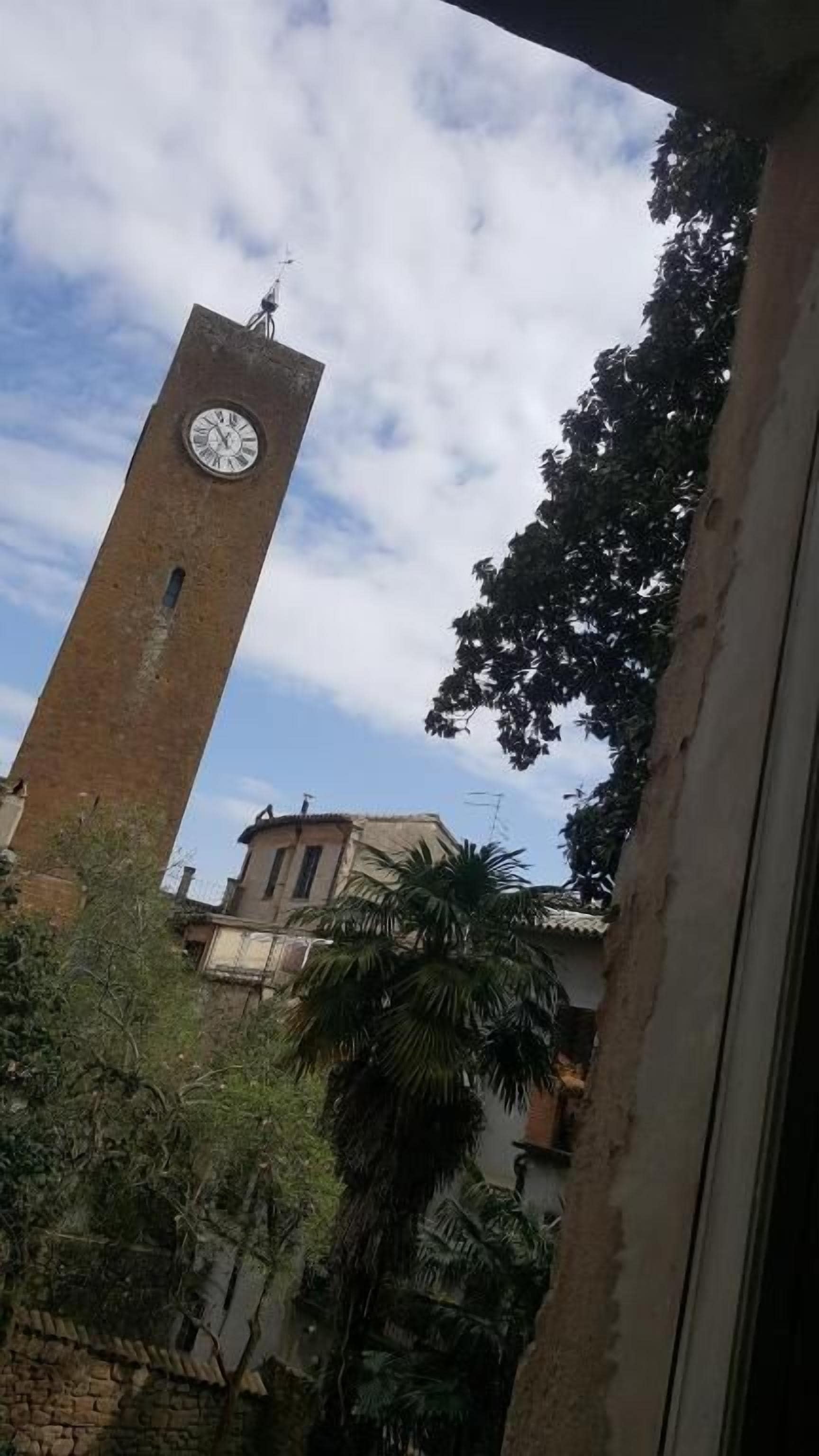Uma torre de tijolos com relógio, rodeada por árvores e edifícios.