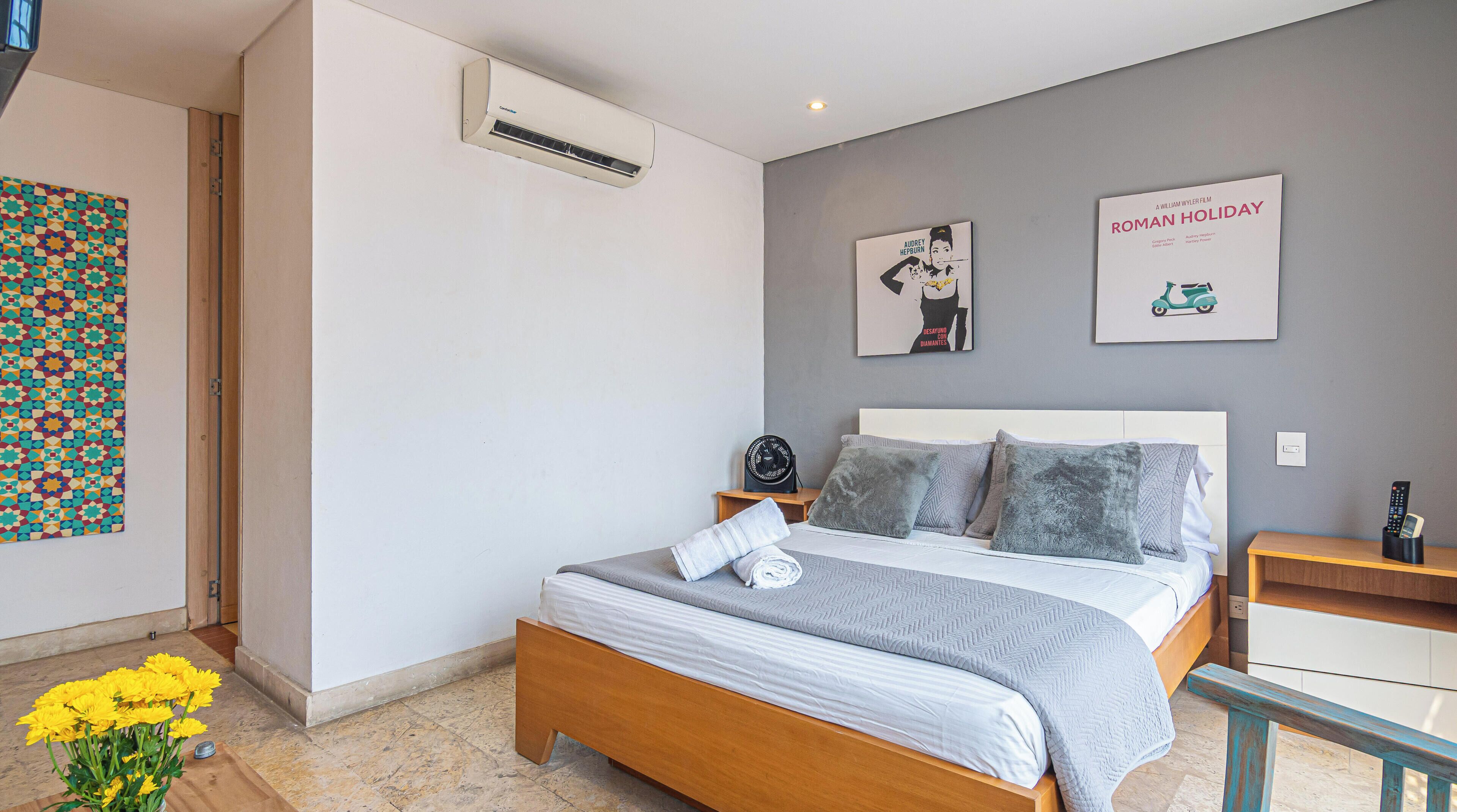 Quarto com cama, almofadas, mesa de cabeceira, ar condicionado fixado na parede e duas fotografias emolduradas.