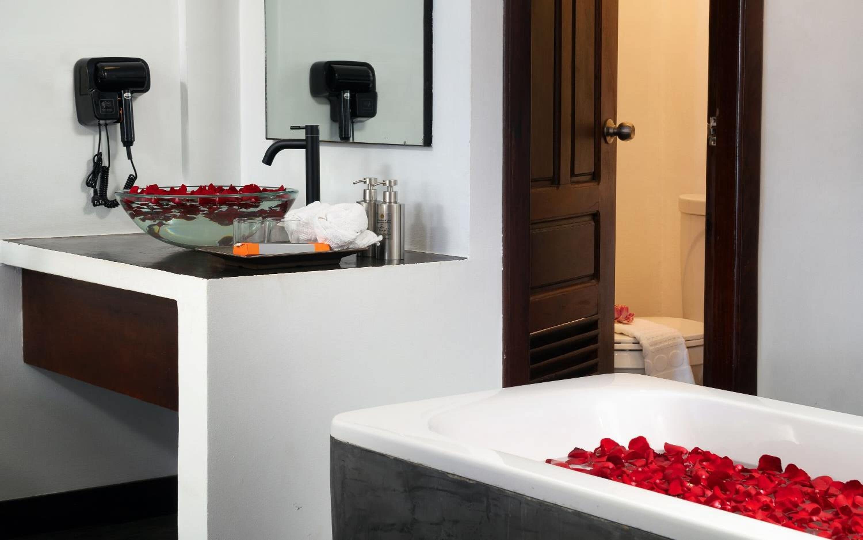 Uma casa de banho com uma pia branca, um espelho, um secador de cabelo preto, uma taça de vidro com pétalas de rosa vermelhas e uma banheira cheia de pétalas de rosa.