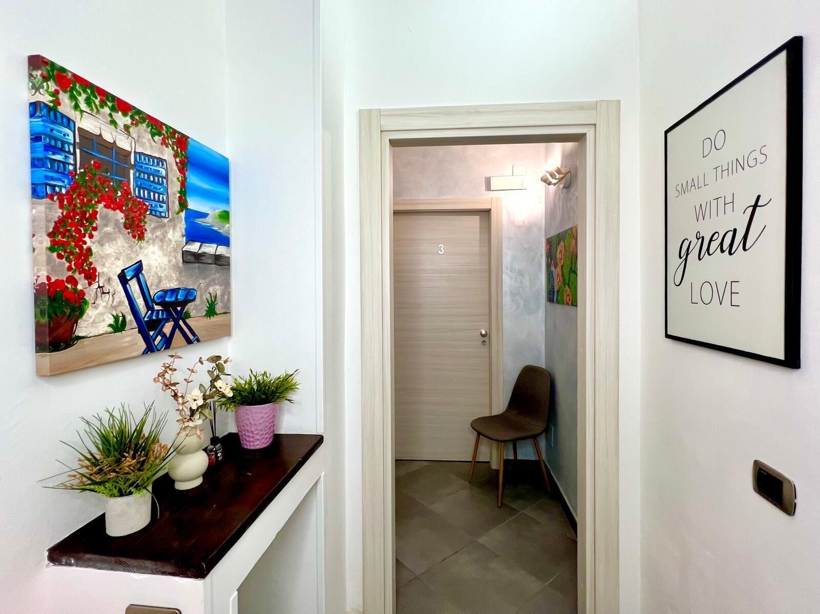 Um corredor com uma porta marcada '3', uma cadeira azul, uma pequena mesa com plantas e uma frase motivacional na parede.