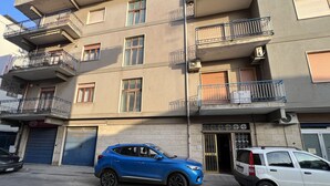 Um edifício com vários andares e varandas, com um carro azul estacionado na frente e um carro branco estacionado ao lado.