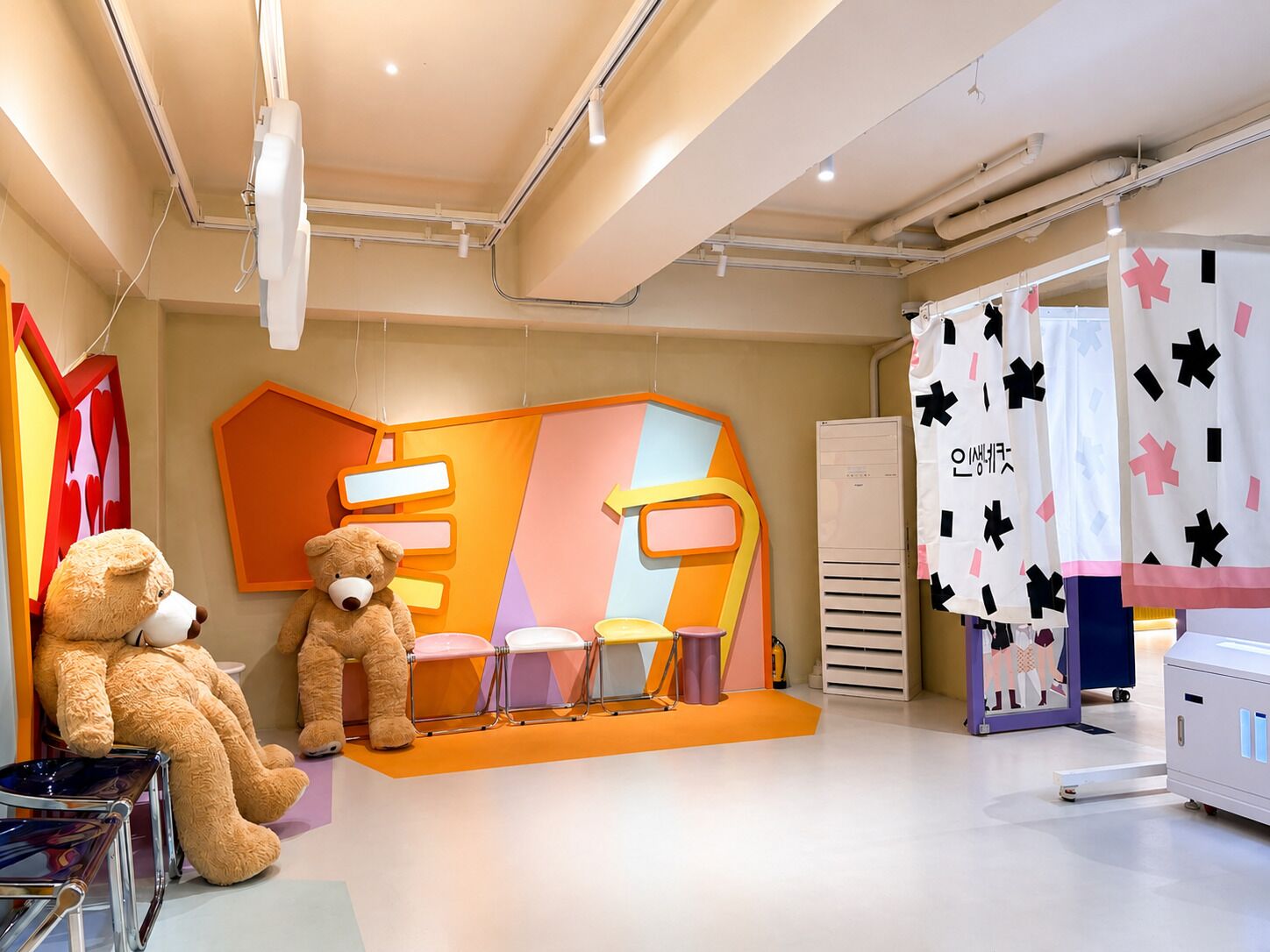 Uma área de recreação infantil com mobiliário colorido, um grande urso de pelúcia e um armário branco.