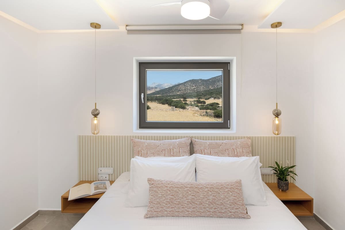 Quarto com cama, mesas de cabeceira, janela com vista para as montanhas e ventilador de teto.