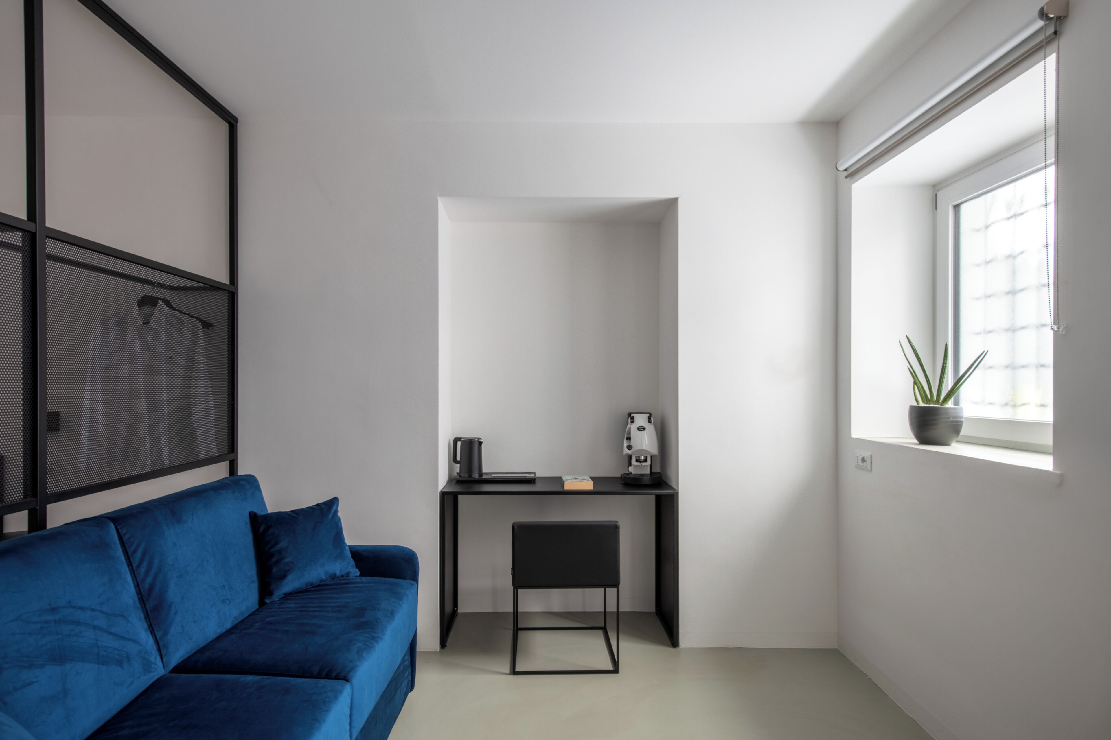 Quarto moderno com sofá azul, escrivaninha com cadeira, planta em vaso e janela com persianas.