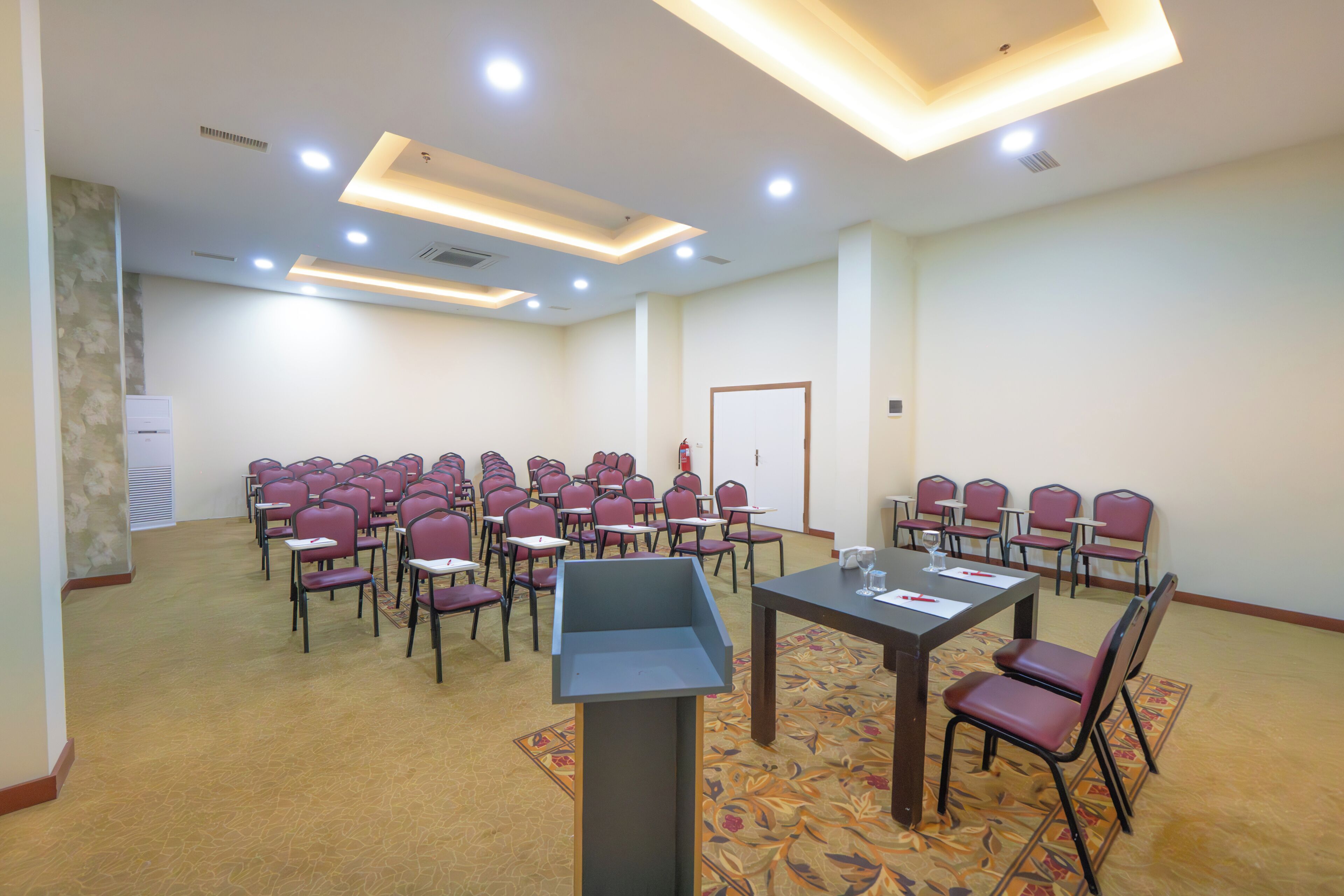 Uma sala de conferência com fileiras de cadeiras, uma mesa com um microfone e piso coberto de carpete.