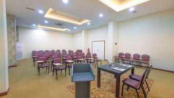 Uma sala de conferência com fileiras de cadeiras, uma mesa com um microfone e piso coberto de carpete.