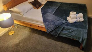 Uma cama com cabeceira de madeira, travesseiro branco, manta escura e duas toalhas enroladas.