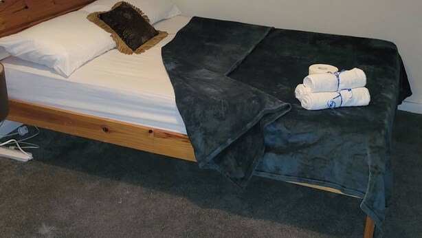 Uma cama de solteiro com cabeceira de madeira, roupa de cama branca, um cobertor escuro e uma toalha dobrada na peseira.