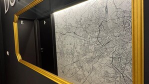 Parede com um espelho grande refletindo um mapa, parede preta com a palavra 'DOMUS' em letras brancas, e uma porta com o número '3'.