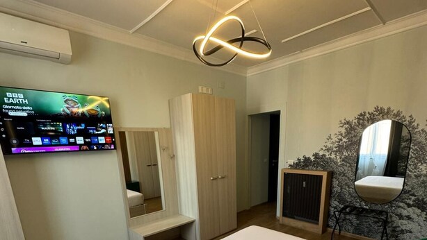 Quarto de hotel com cama, televisão presa na parede, guarda-roupa, espelho e um pequeno espaço de estar.