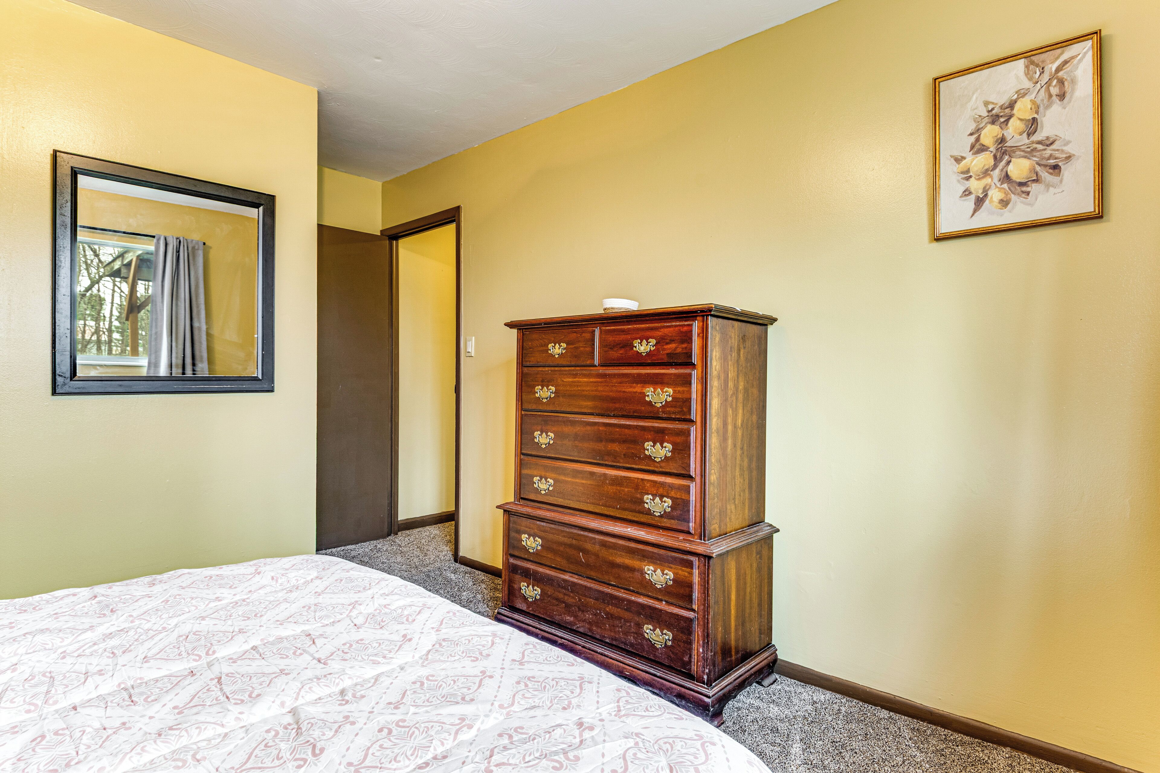 Um quarto com uma cama, uma cômoda de madeira, um quadro pendurado na parede e uma porta que dá para outro cômodo.