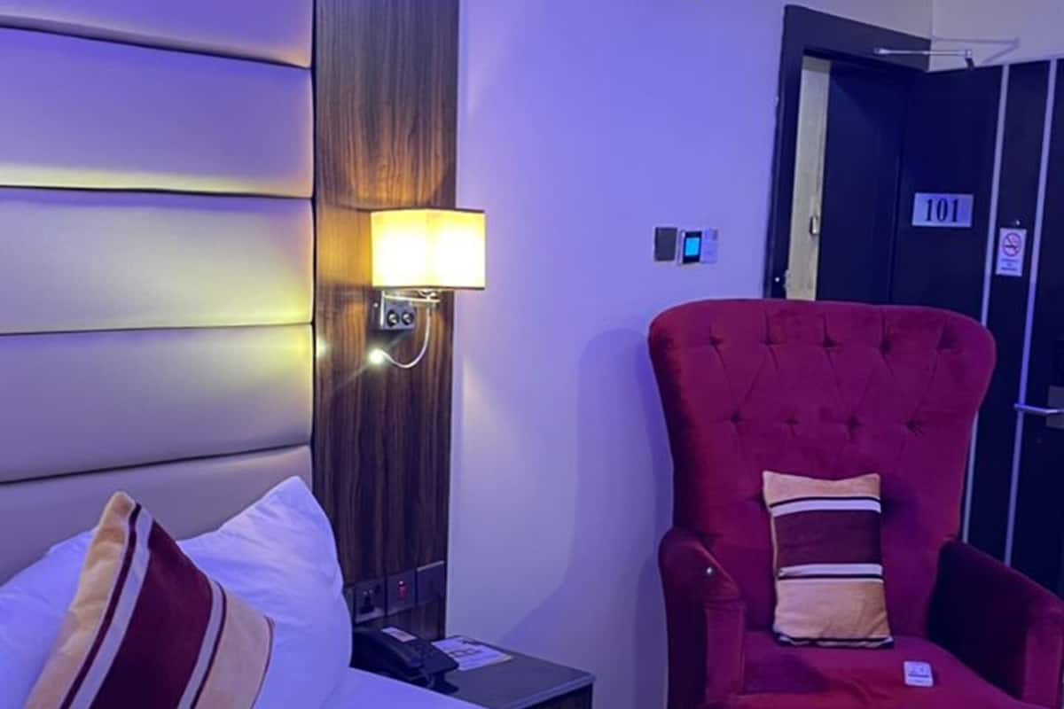 Quarto de hotel com cama, poltrona vermelha, mesa de cabeceira, abajur e porta com a inscrição '101'.