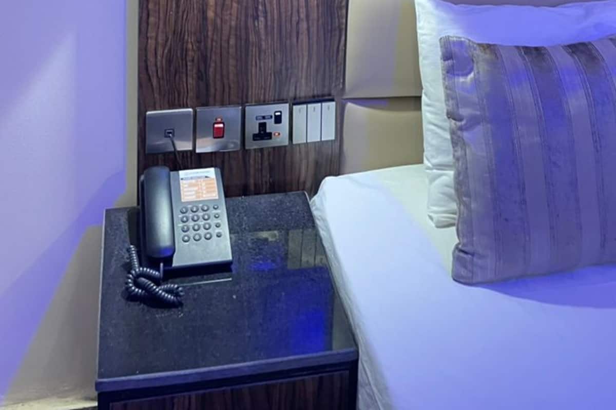 Quarto de hotel com cabeceira de madeira, telefone ao lado da cama, painel de controle e cama com almofada listrada.