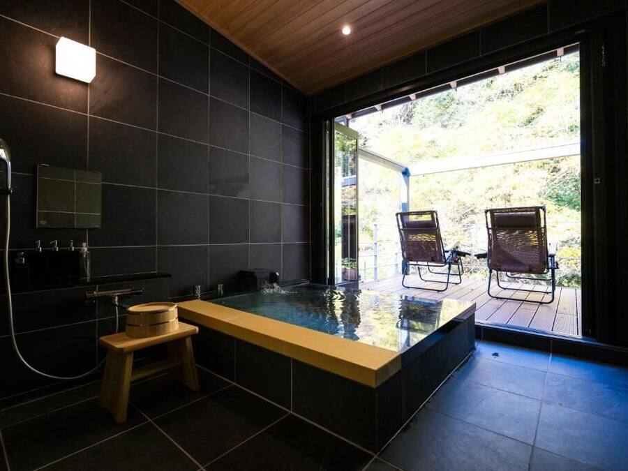 Casa de banho moderna com uma banheira grande, um banquinho de madeira e vista para cadeiras ao ar livre através de uma porta de vidro.