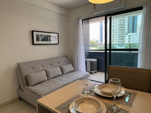 Uma sala de estar moderna com um sofá cinzento, uma mesa de jantar posta com pratos e copos, e uma grande janela com vista para edifícios altos.