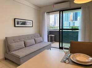 Quarto moderno com sofá cinza, mesa de jantar com taça de vinho e prato, e uma grande janela com vista para edifícios.