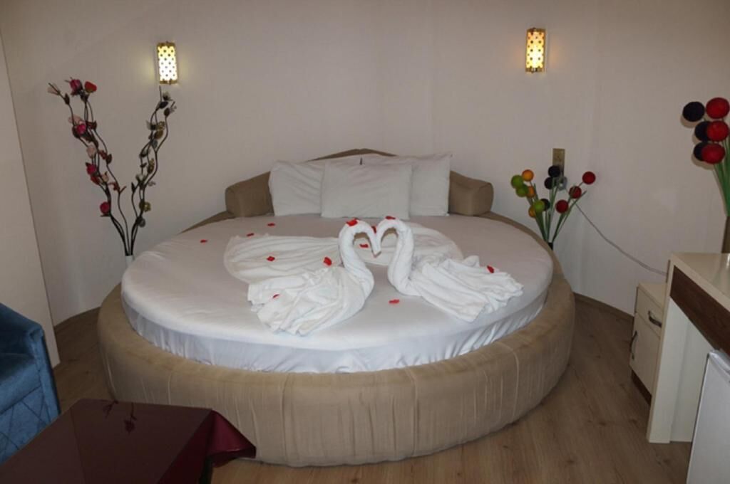 Uma cama redonda com colcha branca, dois toalhas em formato de cisne e pétalas de rosa. Há dois lustres fixados na parede, um vaso com flores e uma pequena mesa com um vaso.