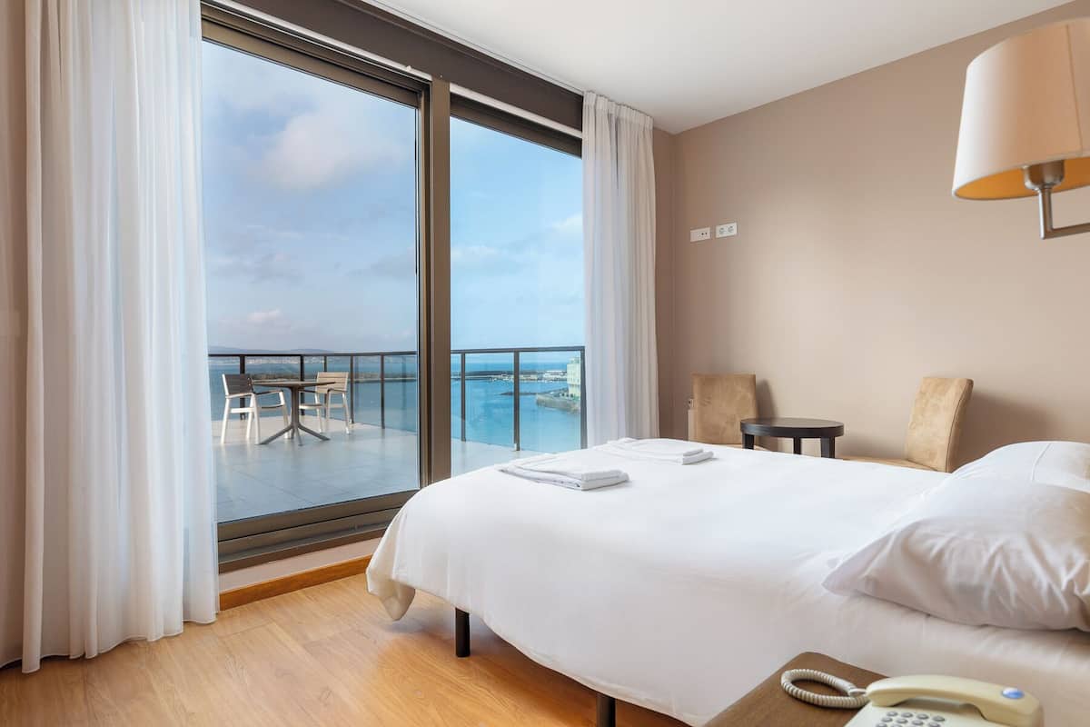 Quarto de hotel com uma cama, mesa de cabeceira com telefone, luminária e vista para o mar através de grandes janelas.