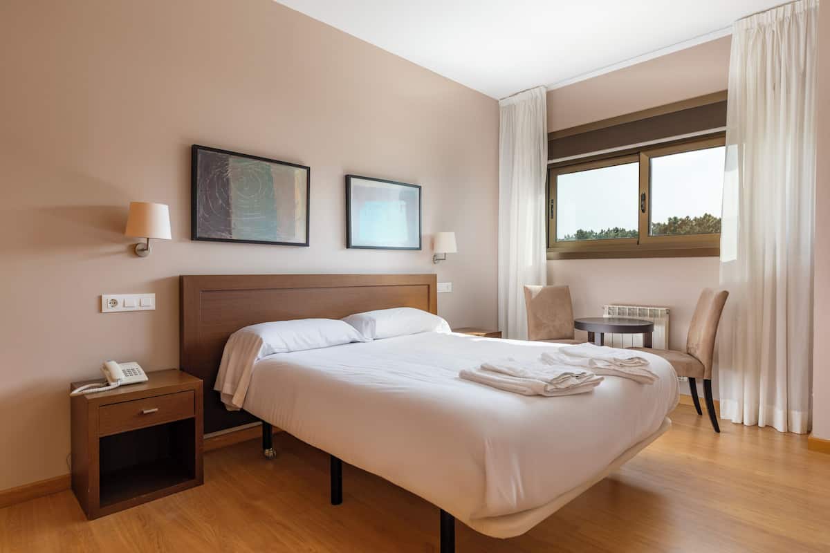 Quarto de hotel com uma cama grande, mesas de cabeceira, uma cadeira, uma pequena mesa e uma janela com cortinas.