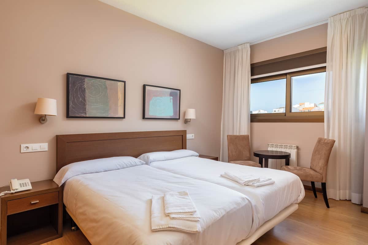 Quarto de hotel com uma cama grande, duas mesas de cabeceira, uma cadeira, uma mesinha e uma janela com cortinas.