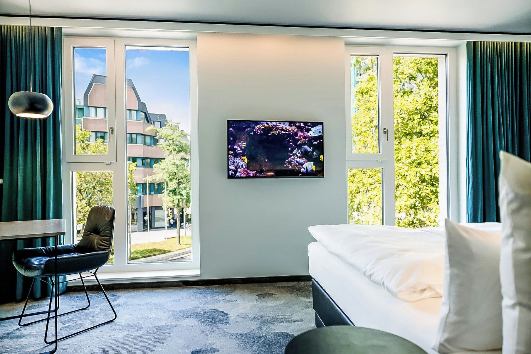 Quarto de hotel com cama, escrivaninha, poltrona, televisão na parede e amplas janelas com cortinas.