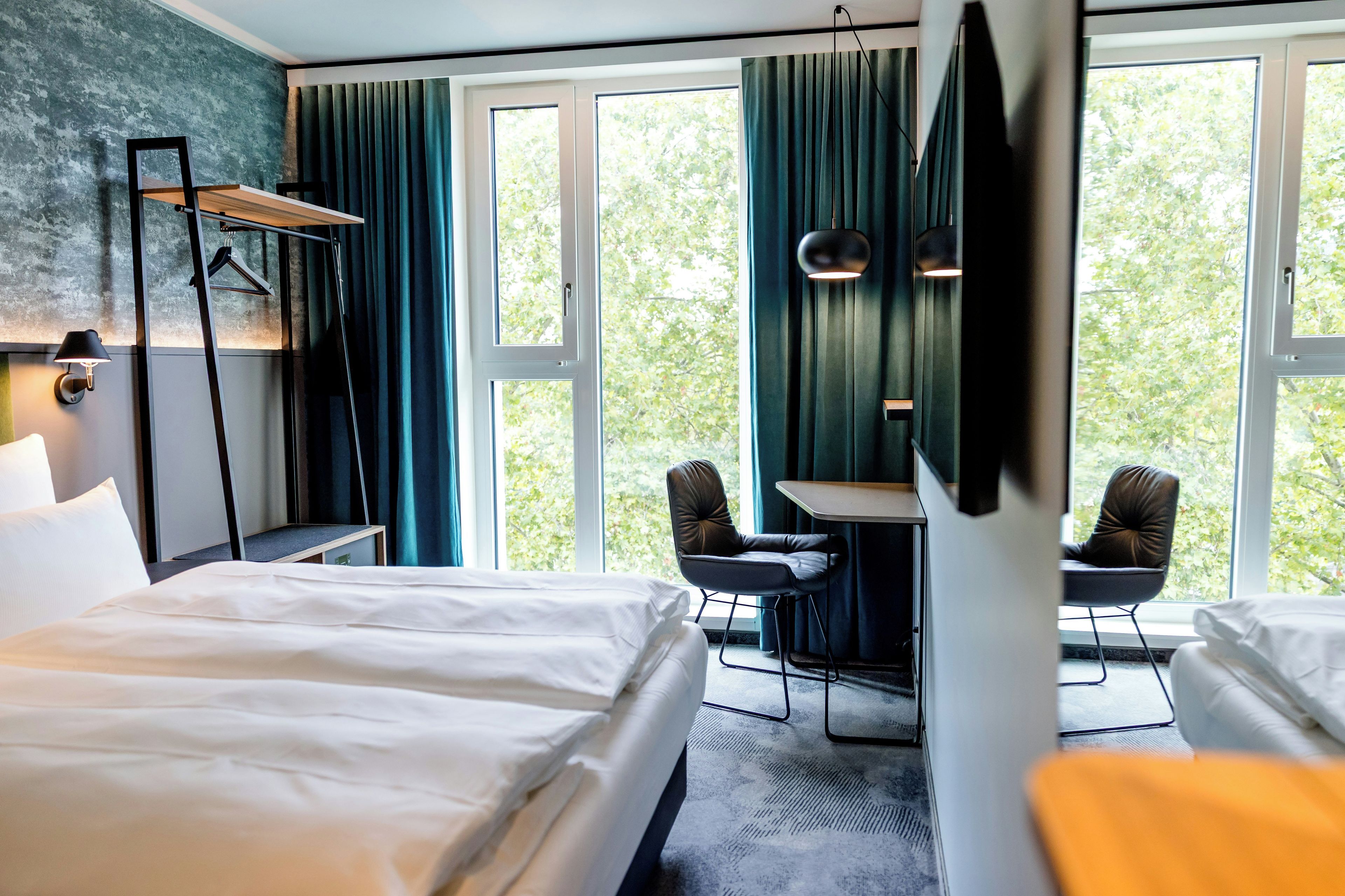 Quarto de hotel com cama, escrivaninha, duas cadeiras, televisão e janelas amplas com cortinas.