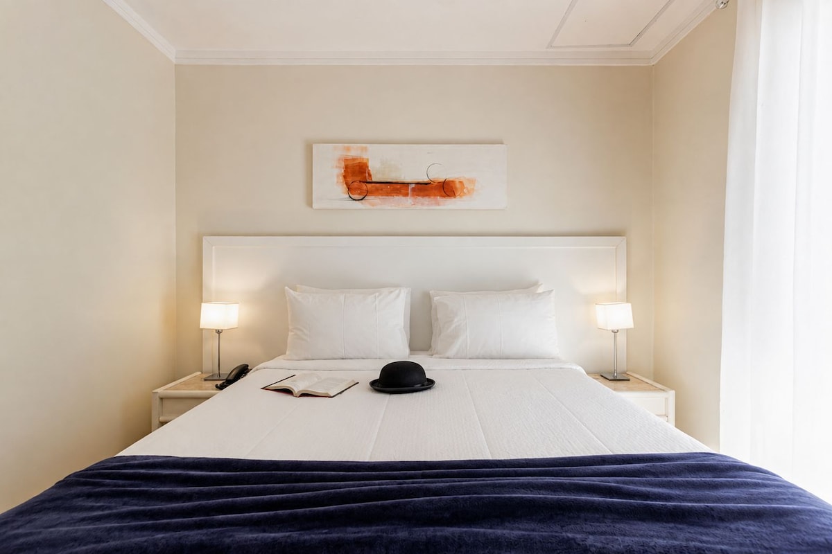 Quarto de hotel com uma cama grande, duas luminárias de cabeceira, um livro, um chapéu e um quadro na parede.