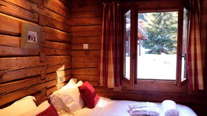 Quarto de cabana de madeira com cama, travesseiros, janela com cortinas e um quadro na parede.