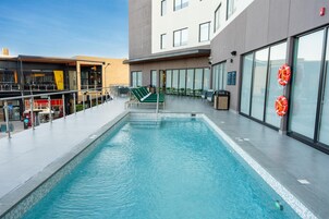 Área de piscina no terraço, com piscina azul cristalina, espreguiçadeiras verdes e um edifício moderno com amplas janelas.