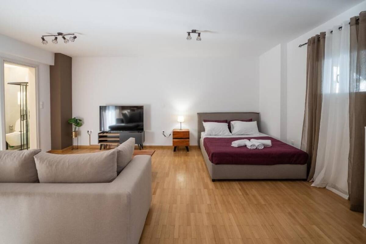 Quarto de hotel moderno com sofá, cama com manta vermelha, televisão, mesa de cabeceira com abajur e piso de madeira.