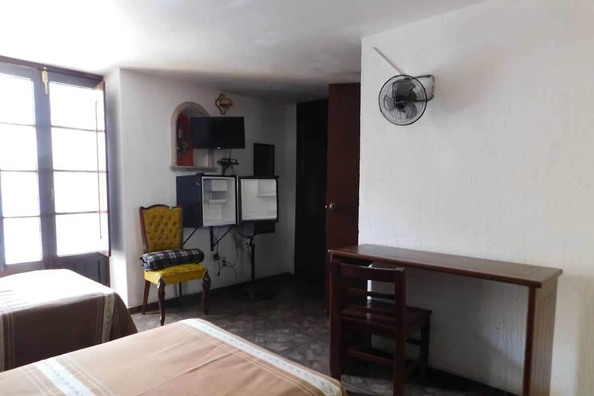 Quarto com cama, escrivaninha, cadeira, ventoinha e televisão.