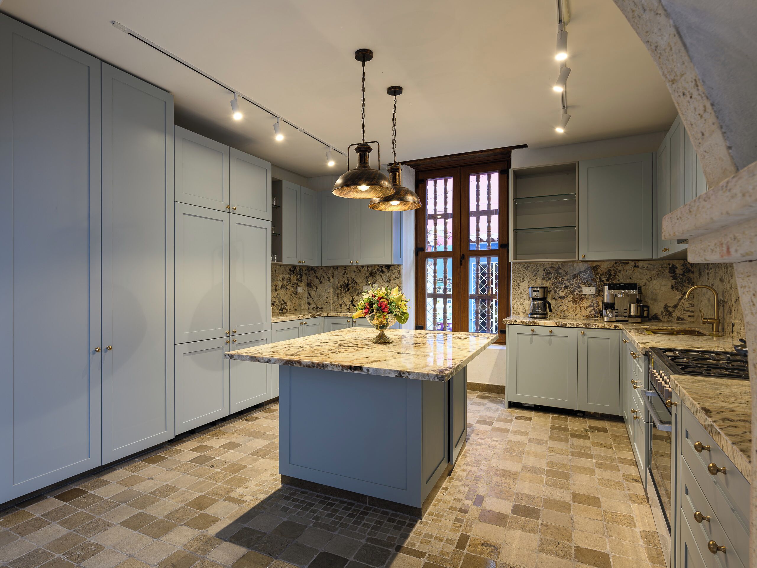 Uma cozinha com ilha central, armários azul-claros, bancada de mármore e luminária pendente.