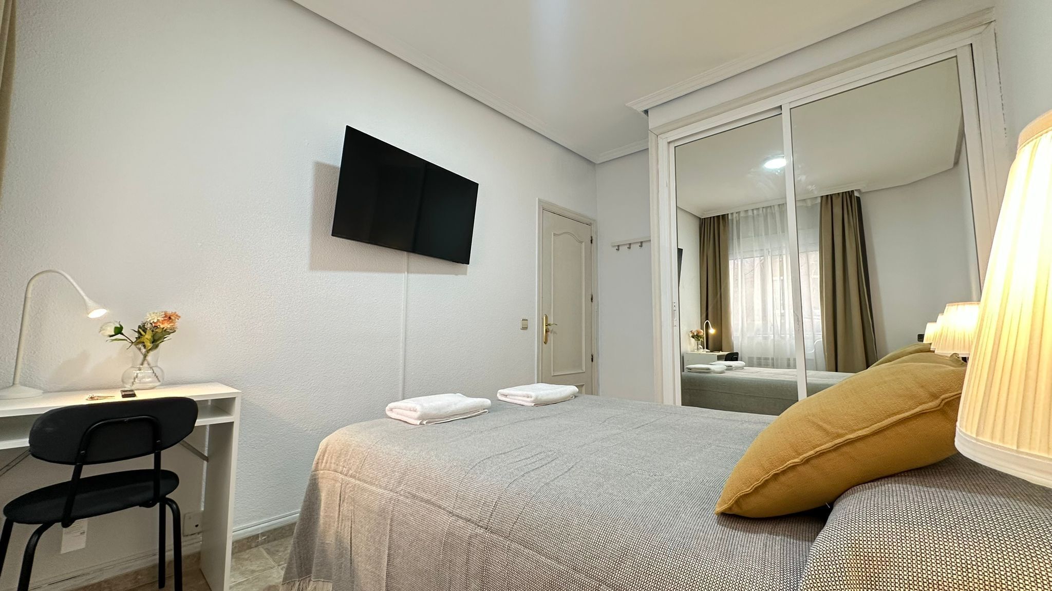 Quarto de hotel com cama, escrivaninha com cadeira, televisão na parede e espelho.