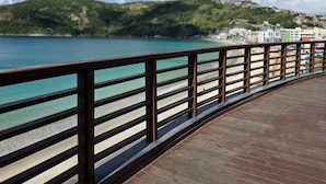 Um calçadão de madeira com corrimão, que leva a uma praia de águas turquesas cristalinas e uma paisagem montanhosa ao fundo.