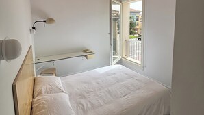 Quarto com teto de madeira, cama com roupa de cama branca, mesa de cabeceira, luminária e janela com vista para edifícios.
