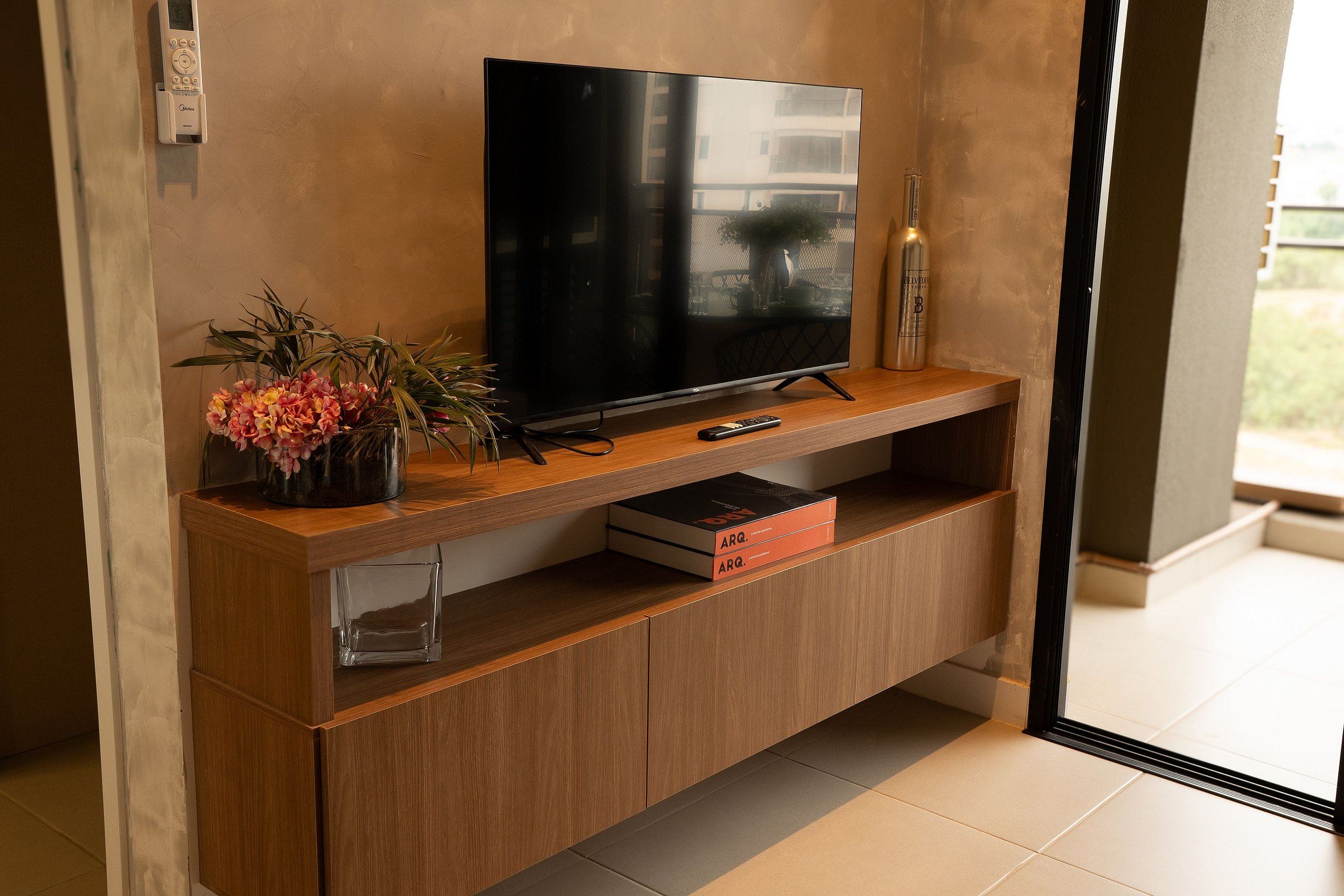 Uma sala de estar moderna com uma TV de tela plana sobre um console de madeira, um controle remoto, um vaso decorativo com flores e uma garrafa.